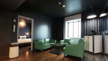 Quarto de hotel moderno com cama, sofás verdes, mesa de centro e escrivaninha com abajur.