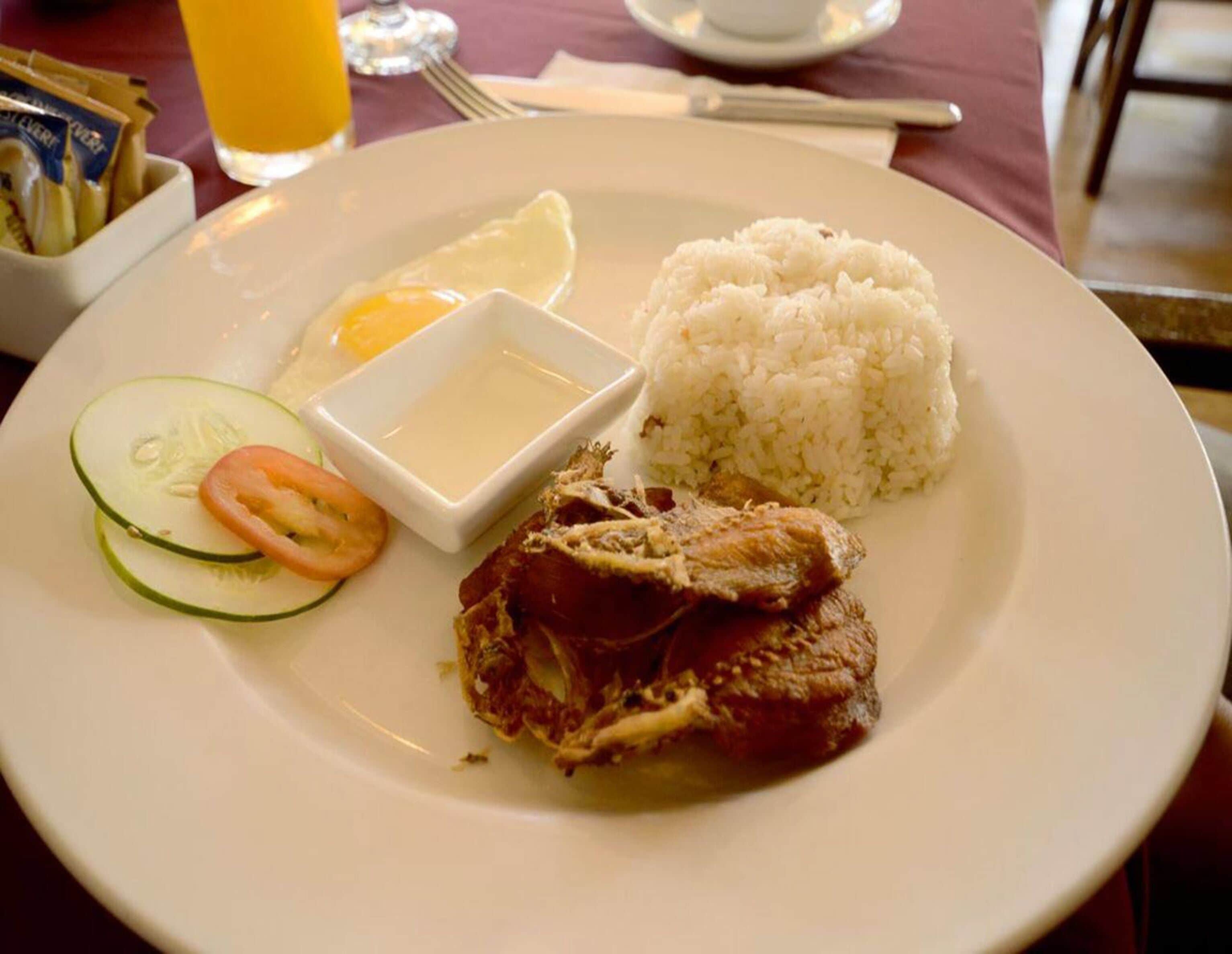 Um prato com arroz, um pedaço de carne, uma fatia de limão, uma pequena tigela de molho e uma fatia de pepino.
