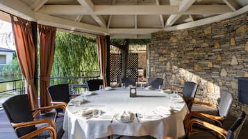 Uma sala de jantar com uma mesa redonda posta para seis pessoas, rodeada por cadeiras de vime, com uma parede de pedra e vegetação ao fundo.