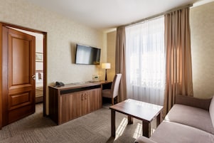 Quarto de hotel com cama, escrivaninha com telefone, TV de tela plana, abajur, poltrona, sofá e janela com cortinas.
