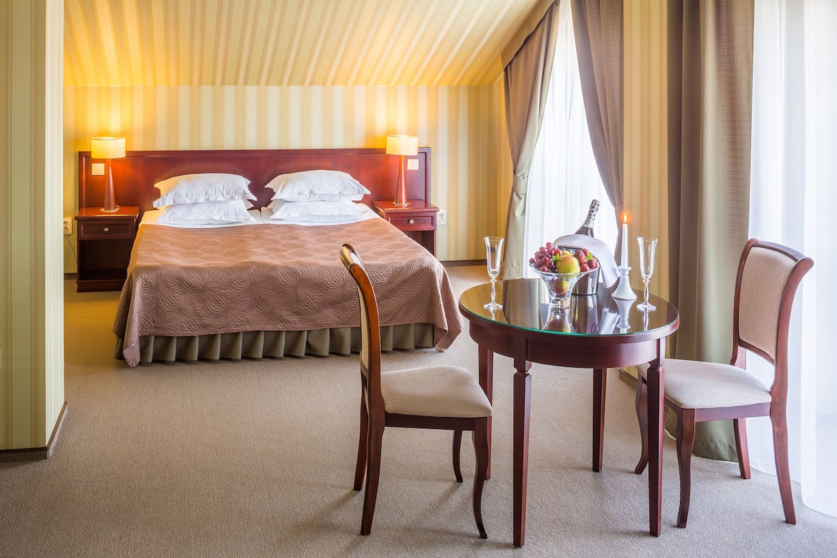 Quarto de hotel com uma cama grande, duas mesas de cabeceira com abajures, uma mesa de jantar redonda com cadeiras e um vaso de frutas.