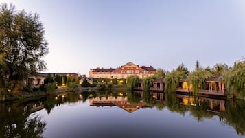 Um grande hotel refletido em águas calmas, rodeado por árvores e uma pequena ponte.