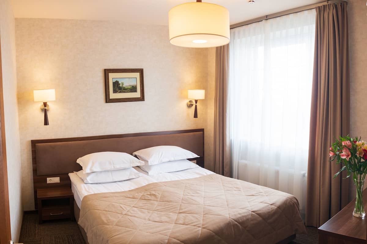Quarto de hotel com uma cama grande, duas luminárias de cabeceira, um quadro na parede e uma janela com cortinas.