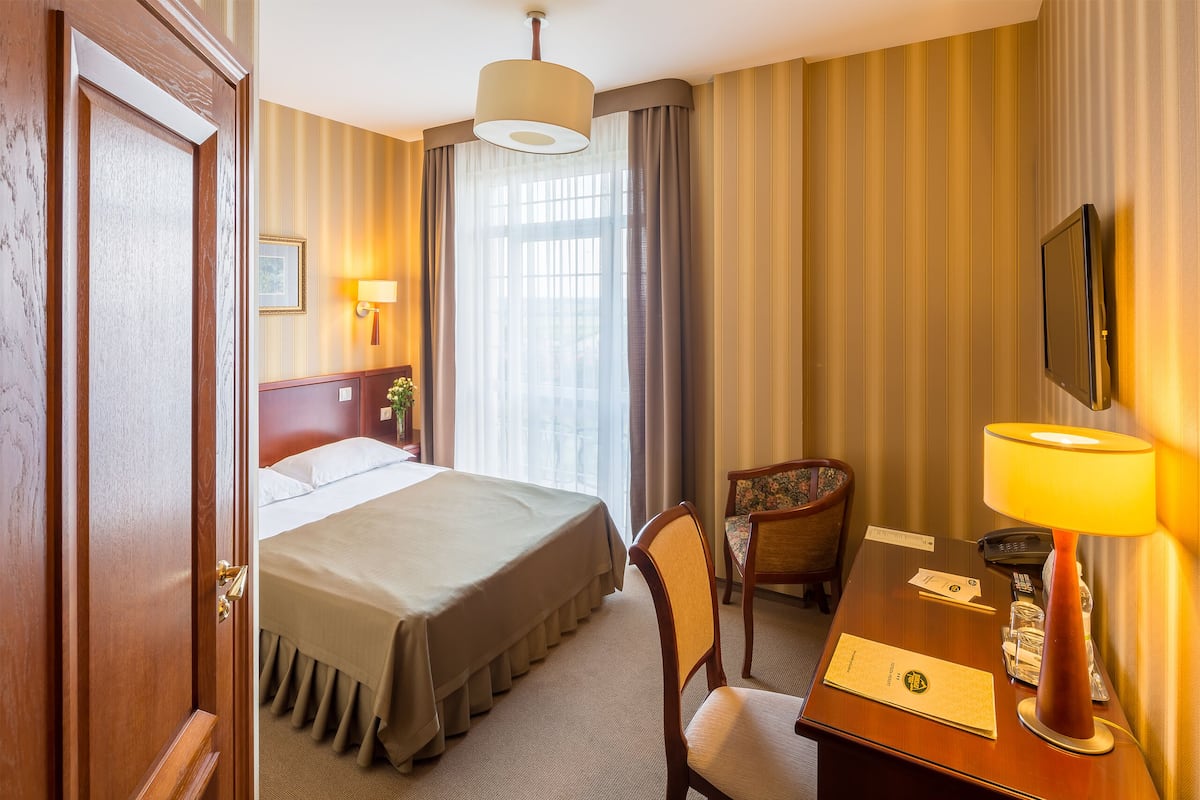 Quarto de hotel com cama, escrivaninha com luminária, cadeira, televisão e janela com cortinas.