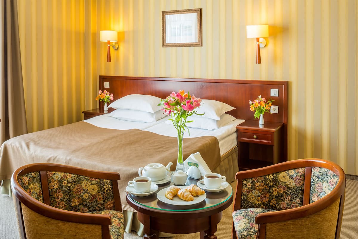 Quarto de hotel com uma cama, duas mesas de cabeceira com luminárias, uma mesinha com serviço de chá e duas cadeiras.