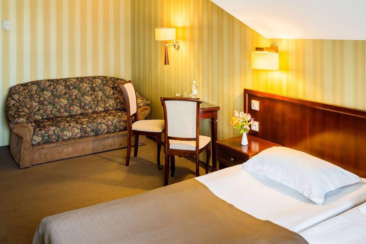 Quarto de hotel com sofá floral, uma escrivaninha de madeira, uma cadeira, cama com roupa de cama branca e duas luminárias fixadas na parede.