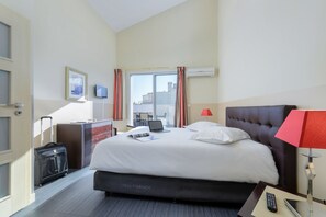 Quarto de hotel com cama, escrivaninha, cadeira, mala, abajur e janela com cortinas.