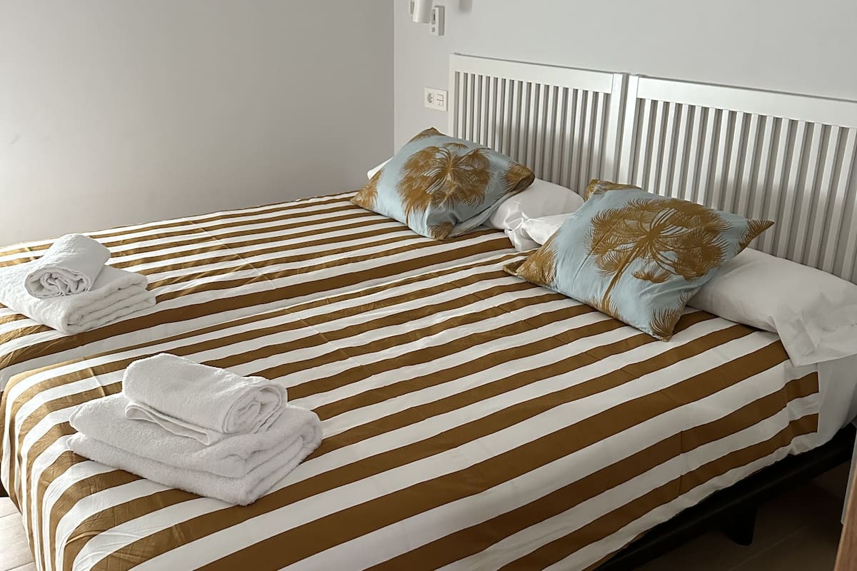Uma cama bem arrumada com roupa de cama listrada, duas almofadas com estampas de folhas de palmeira e um ar-condicionado branco fixado na parede.