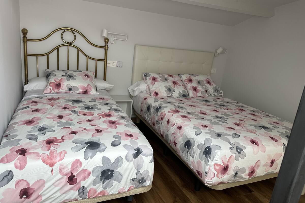 Duas camas de solteiro com roupa de cama floral, uma cabeceira branca e uma mesa de cabeceira com abajur.