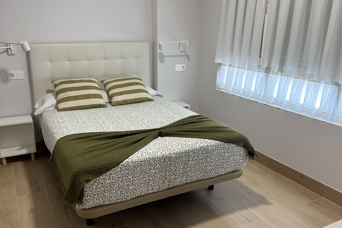 Um quarto com cama, mesa de cabeceira, janela com persianas e luminária fixada na parede.