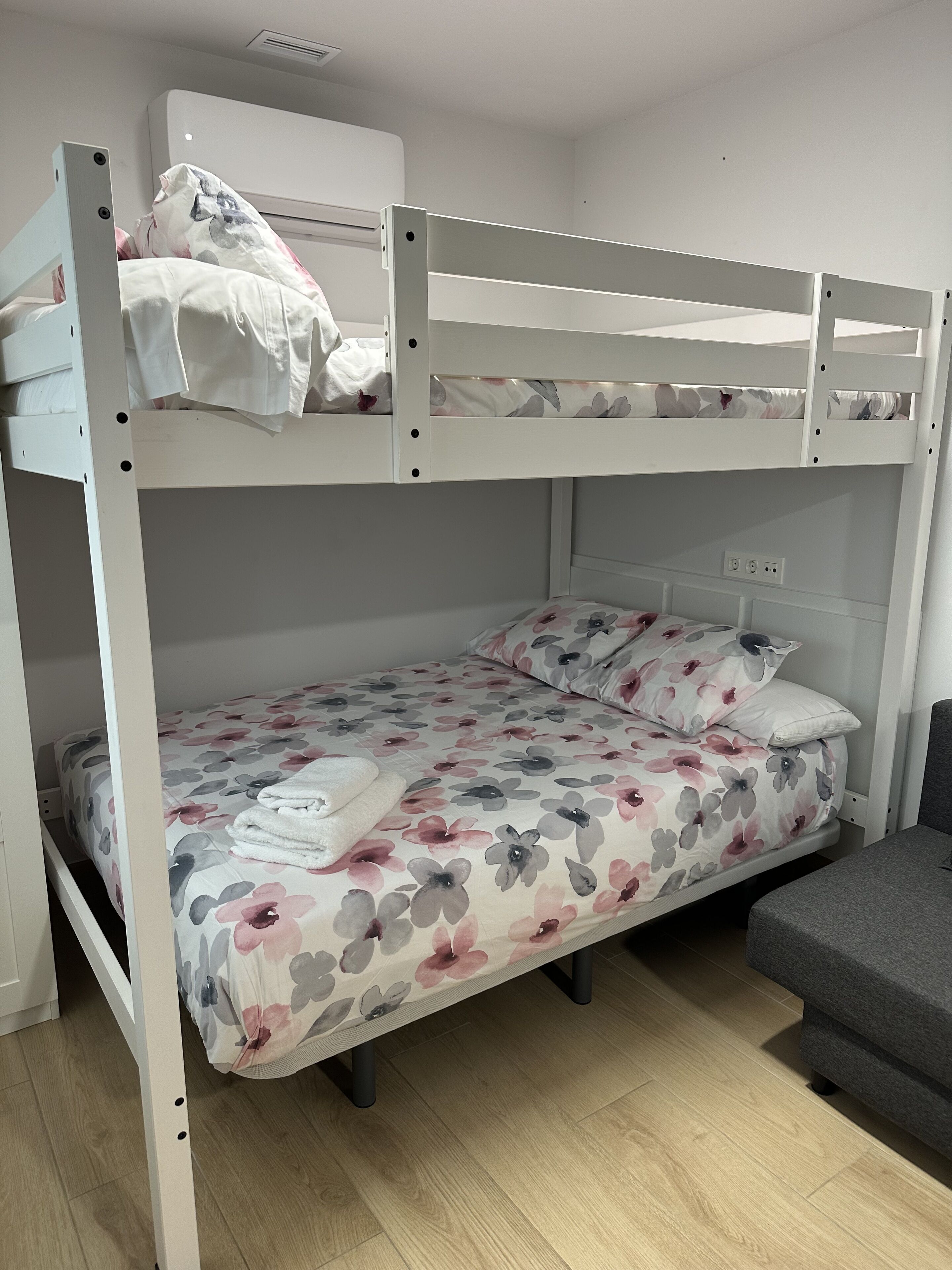 Uma cama beliche com colcha florida e um sofá cinza em um quarto com piso de madeira clara.