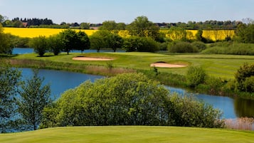 Um campo de golfe com um lago, árvores e um campo amarelo ao fundo.
