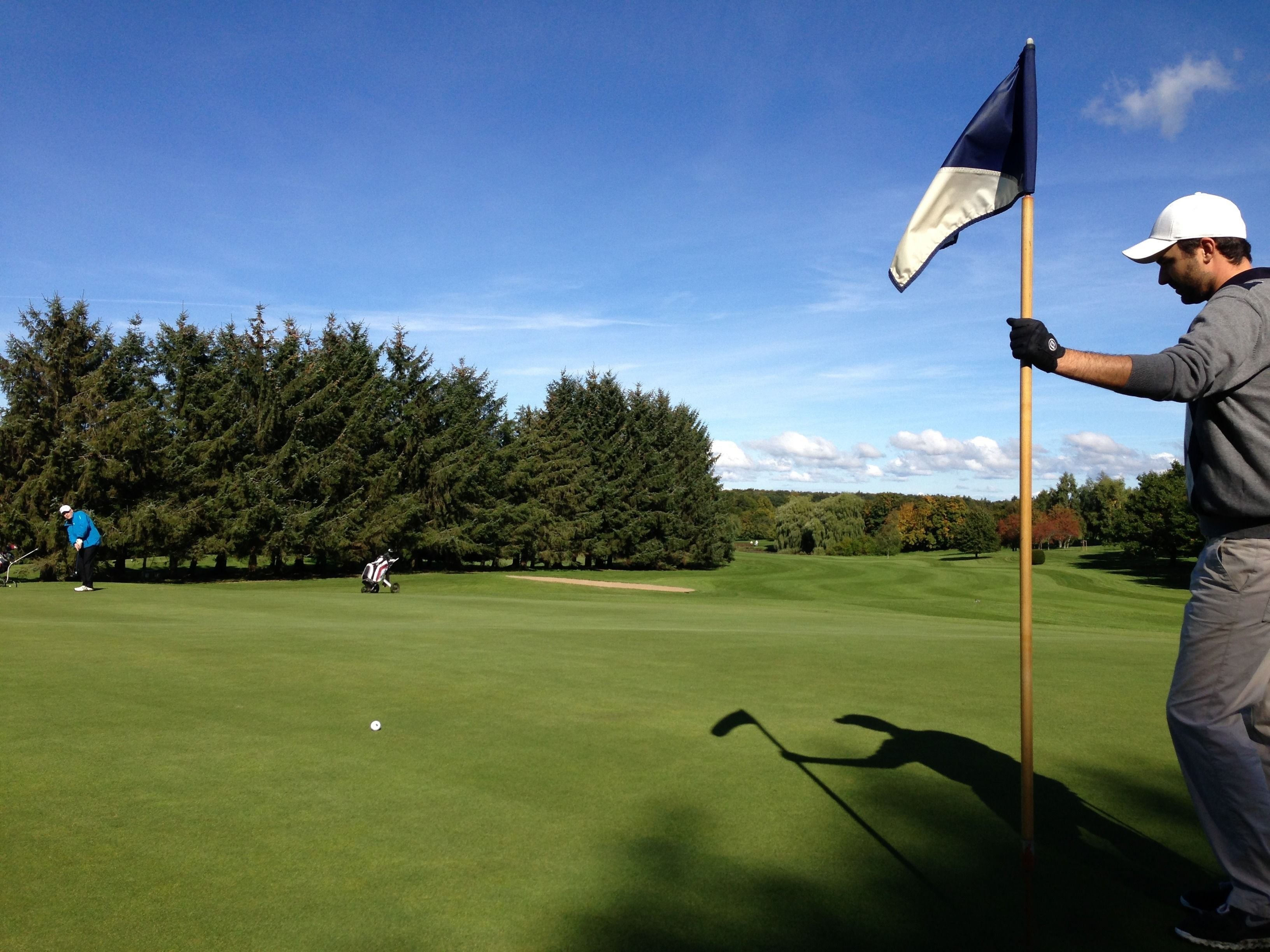 Um campo de golfe com haste de sinalização, um jogador de golfe com jaqueta azul e outro jogador à distância.