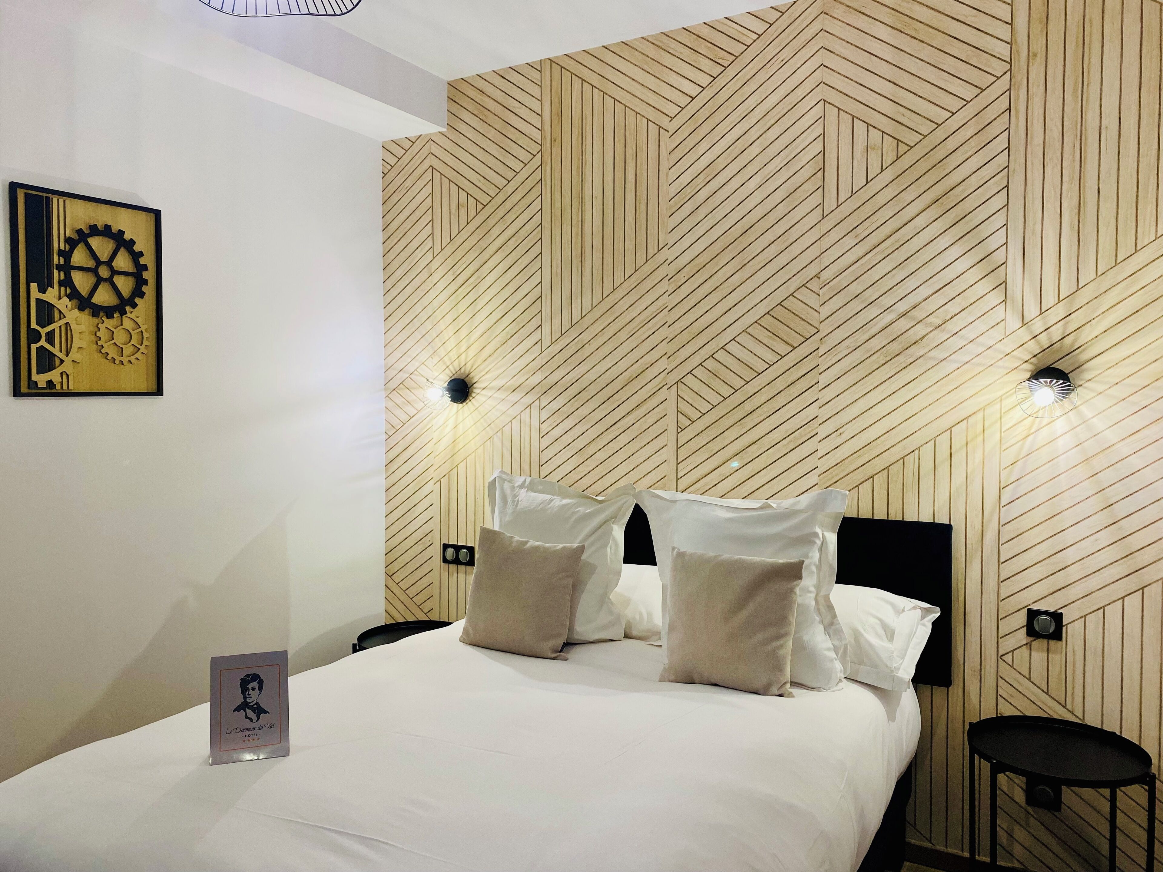 Cama bem arrumada com lençóis brancos e duas almofadas, parede com padrão geométrico de madeira, quadro na parede e dois luminários fixados na parede.