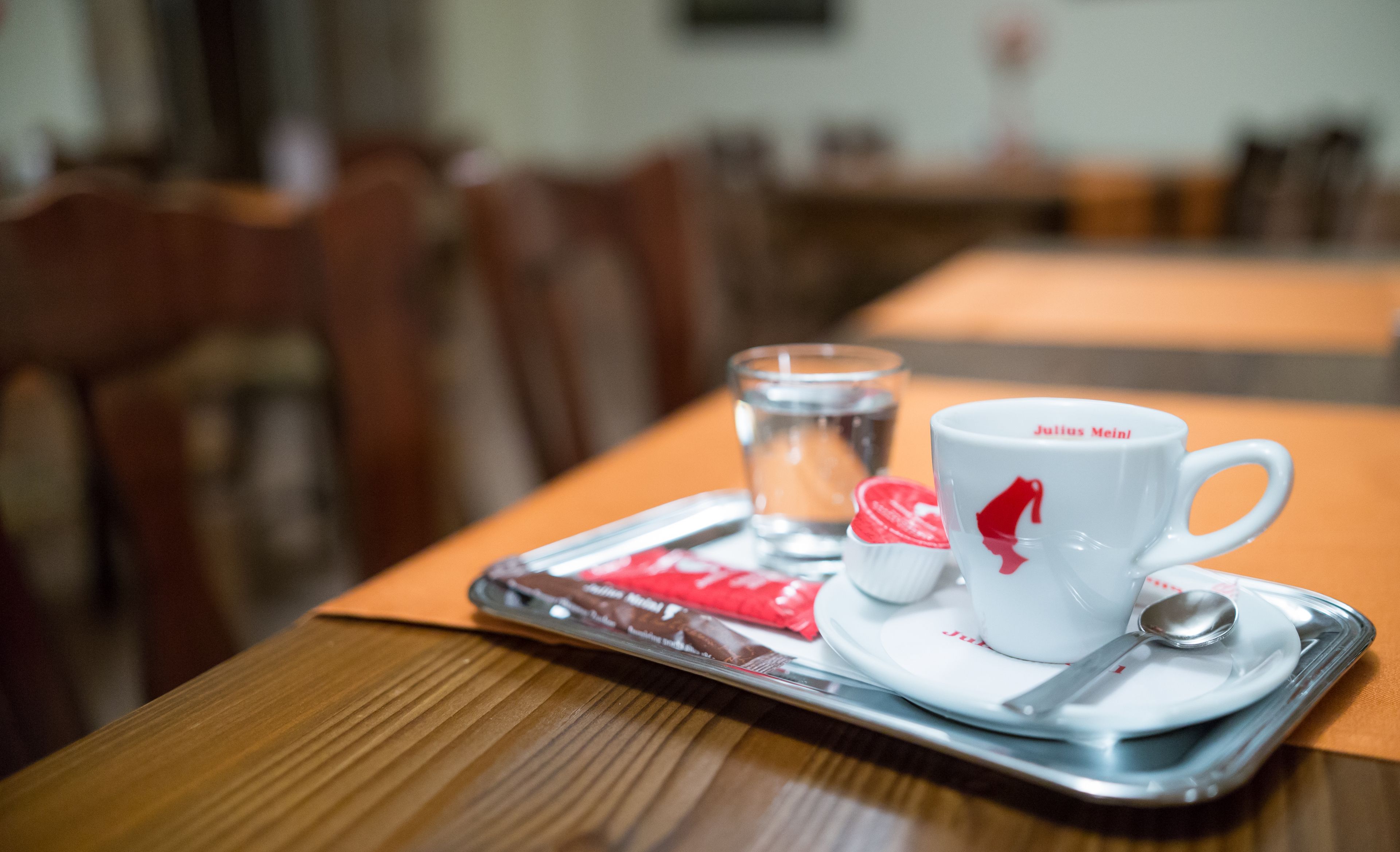 Uma bandeja com uma xícara branca, pires e colher, um copo de água e um guardanapo vermelho sobre uma mesa de madeira em uma sala de jantar.