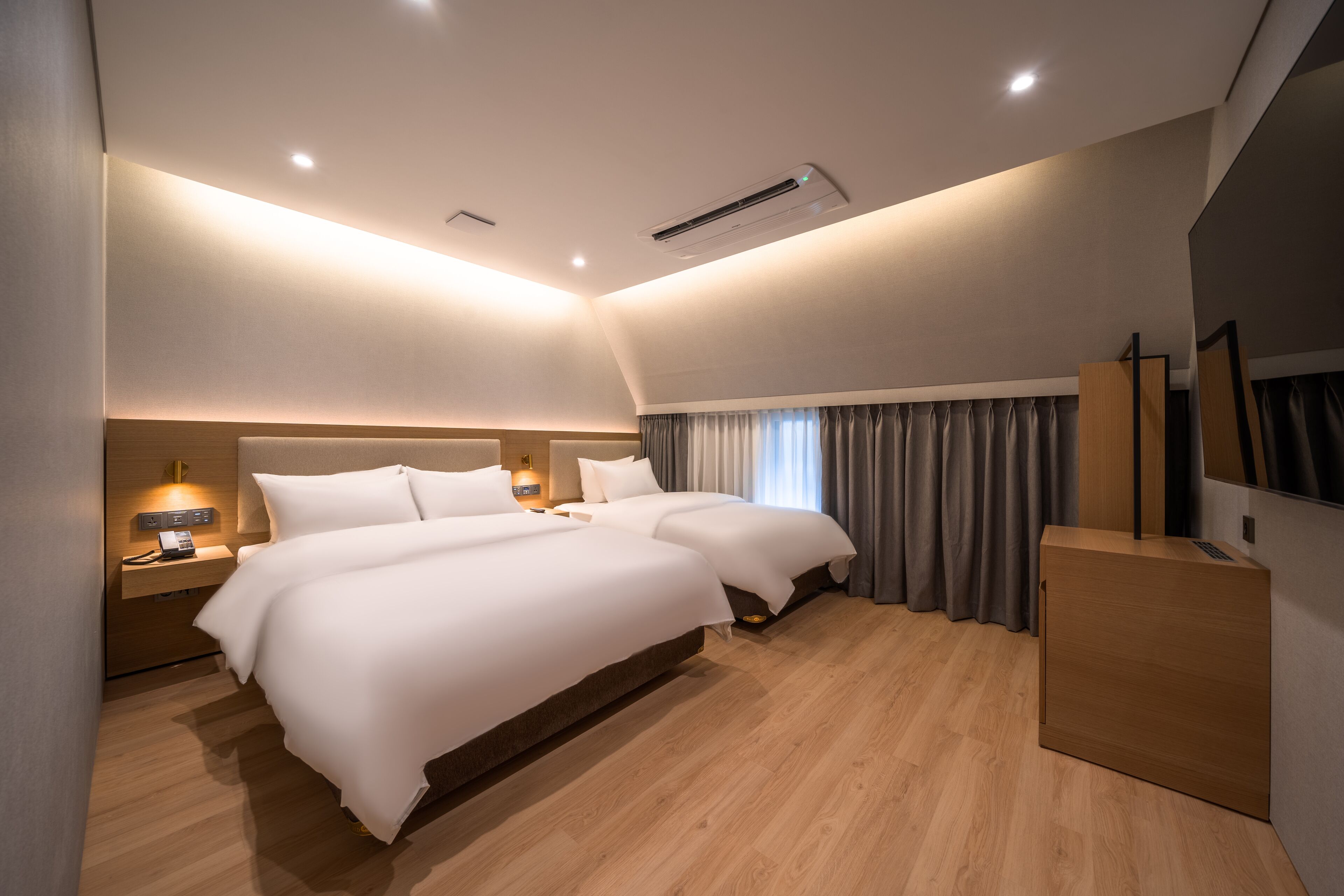 Quarto de hotel moderno com duas camas, piso de madeira, televisão embutida e mesa de cabeceira com abajur.