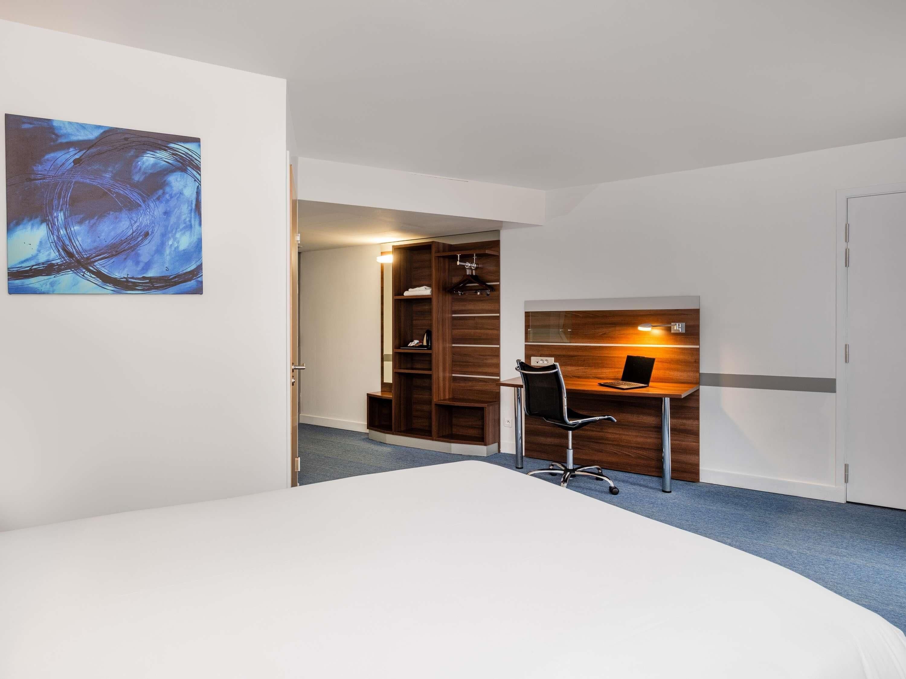 Quarto de hotel com cama, secretária, cadeira, guarda-roupa e um quadro na parede.