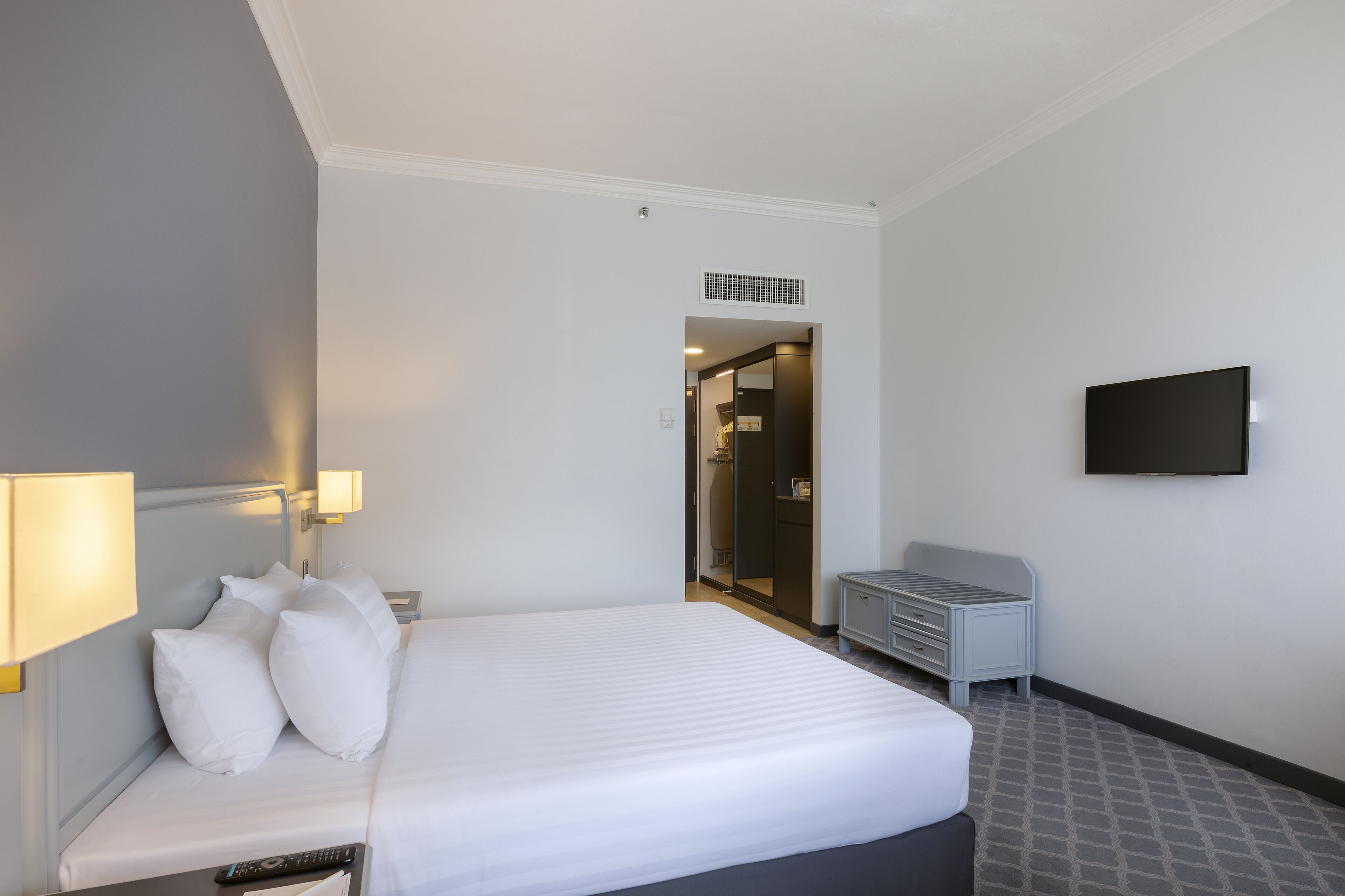 Quarto de hotel com cama, mesa de cabeceira, televisão na parede e guarda-roupa.