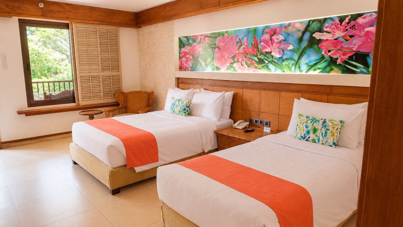Quarto de hotel com duas camas, uma janela grande com persianas, cabeceira de madeira e uma pintura de flores.