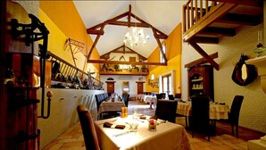 Uma sala de jantar com pé-alto, vigas de madeira aparentes e um candelabro. As mesas estão postas com toalhas e talheres.