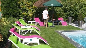 Uma área de piscina com espreguiçadeiras verdes e rosa, um homem caminhando e um guarda-sol verde.