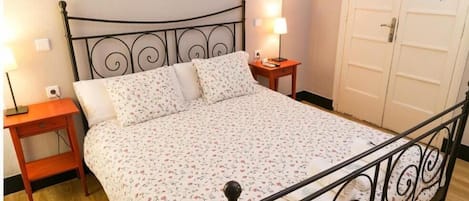 Uma cama de solteiro com colcha florida, uma mesinha de cabeceira com abajur e uma porta com aviso.