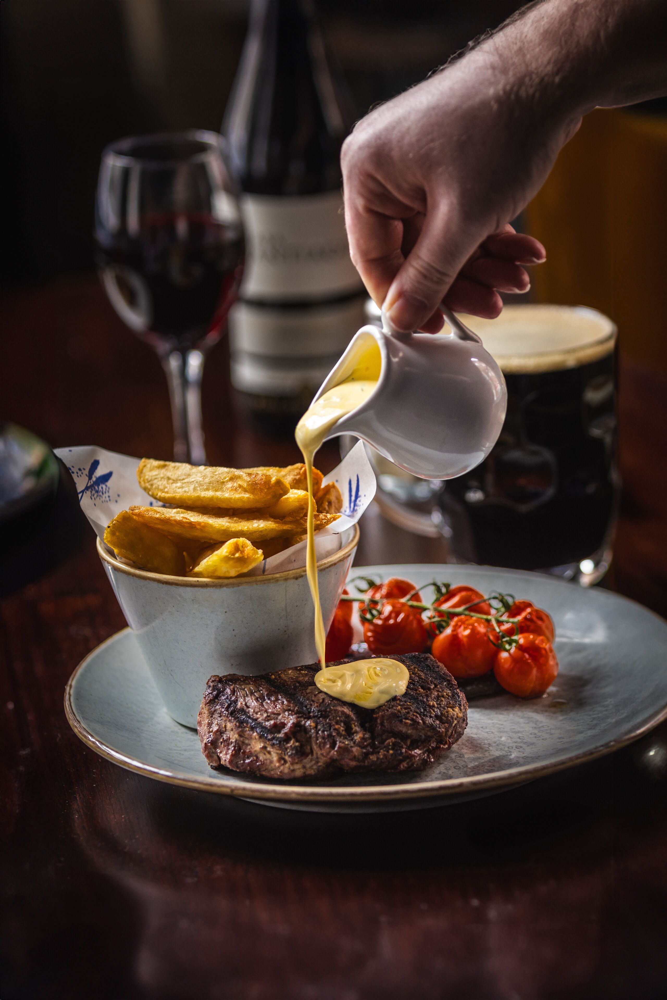 Um prato com bife, batatas fritas e tomates cereja, e uma mão regando as batatas com molho.