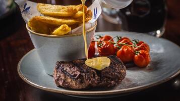 Um prato com bife, batatas fritas e tomates cereja, e uma mão regando as batatas com molho.