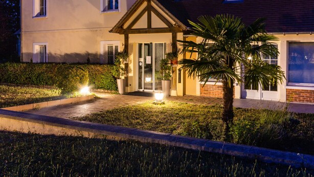 Uma casa de dois andares com uma entrada bem iluminada, um pequeno alpendre e um jardim com uma palmeira.
