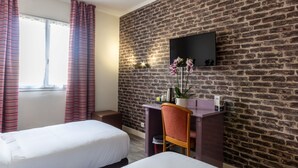 Quarto de hotel com duas camas, uma escrivaninha, uma cadeira, uma TV fixada numa parede de tijolos e uma janela com cortinas.