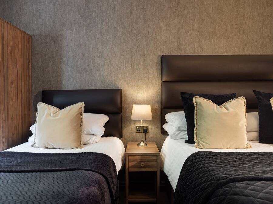 Quarto de hotel com duas camas, cada uma com cabeceira, mesa de cabeceira e luminária, além de um guarda-roupa.