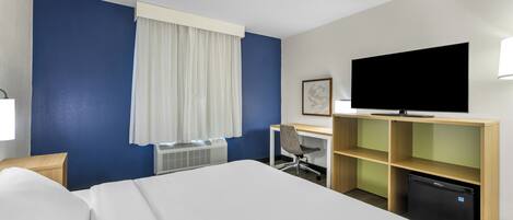 Quarto de hotel com cama, escrivaninha, cadeira, televisão, mini-geladeira e janela com cortinas.