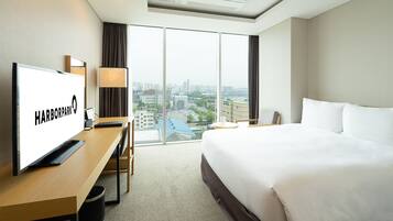 Quarto de hotel com uma cama grande, uma mesa com uma TV exibindo "HARBORPARK", vista para a cidade através de janelas amplas e um design moderno no teto.