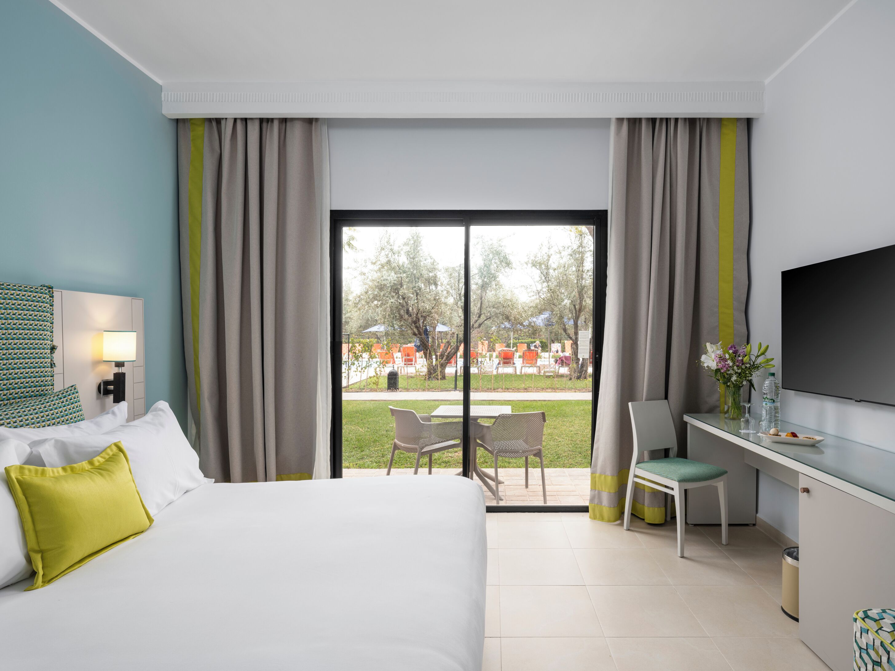 Quarto de hotel com cama, escrivaninha com cadeira, televisão e varanda com área de estar ao ar livre.