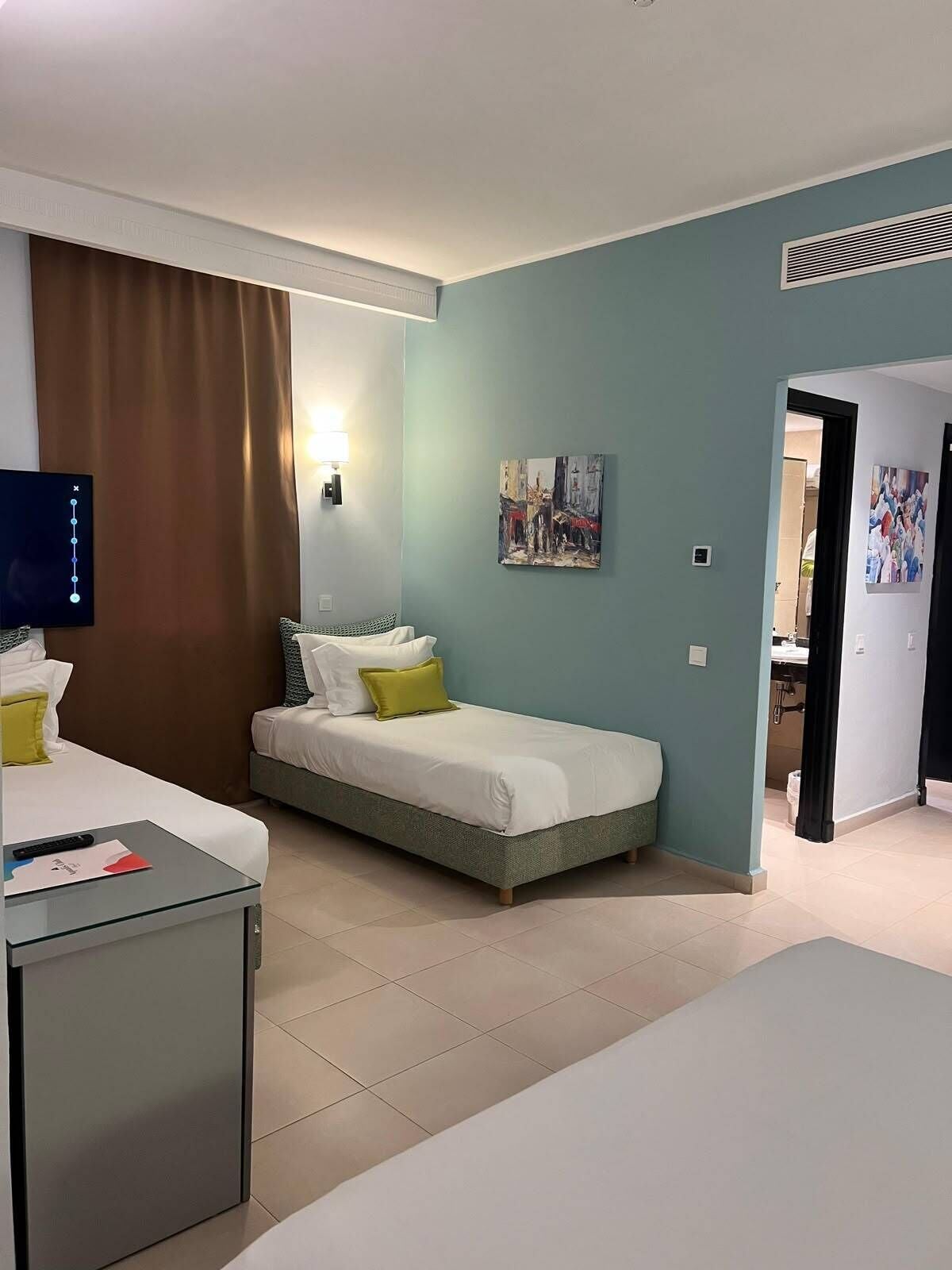 Quarto de hotel com cama, escrivaninha com computador, televisão e obras de arte nas paredes.