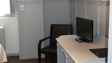 Quarto compacto com cama, escrivaninha com computador, cadeira e pequena cozinha.
