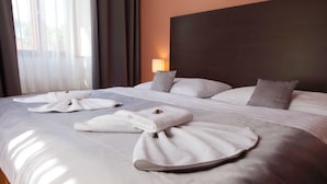 Quarto de hotel com cama grande, duas almofadas, abajur e vista para o exterior através de cortinas translúcidas.