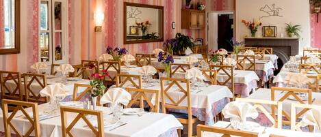 Uma área de refeições com várias mesas preparadas para o serviço, cadeiras de madeira e centros de mesa florais.