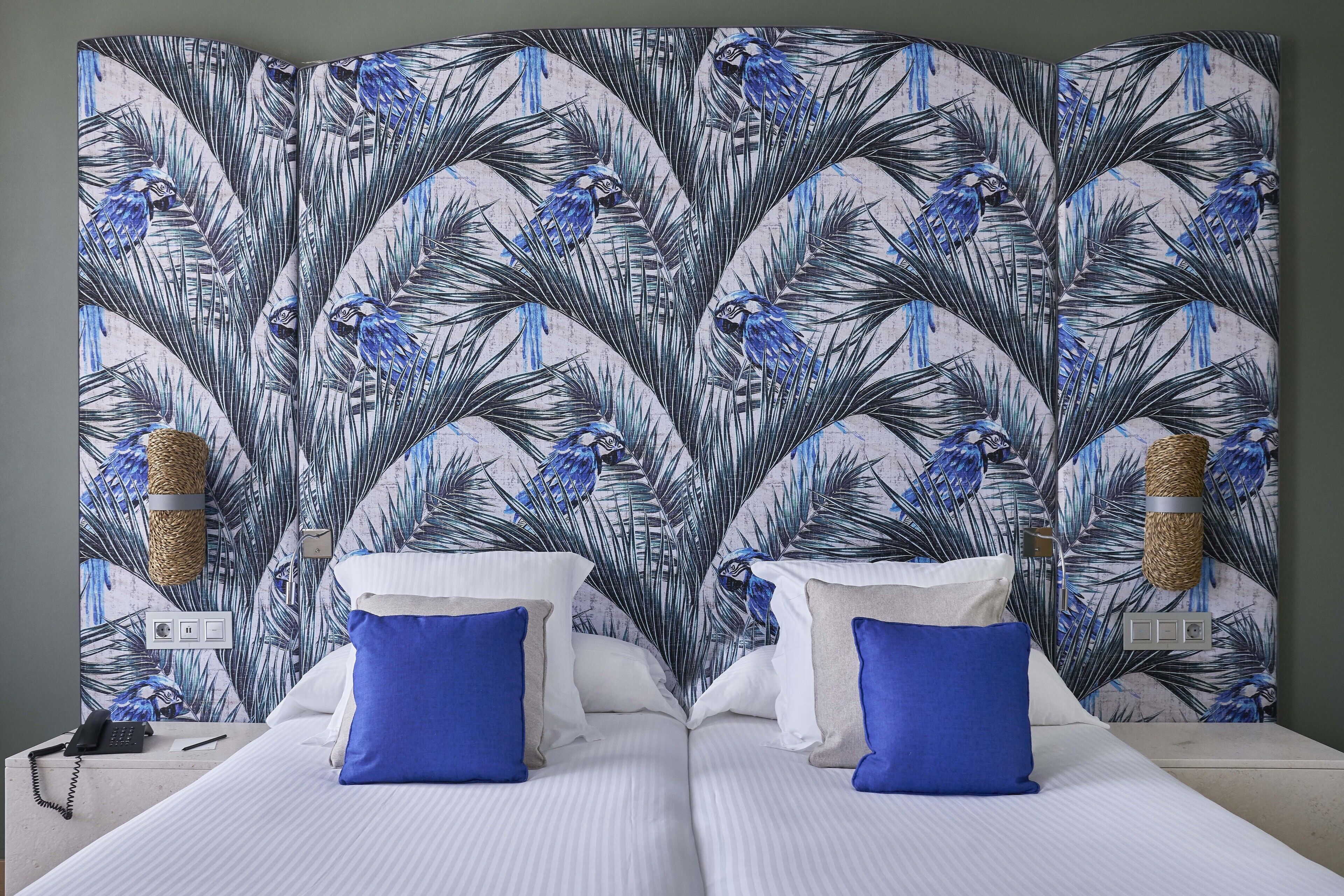 Quarto com duas camas, cada uma com um travesseiro azul, e uma parede com padrão de folhagem tropical.