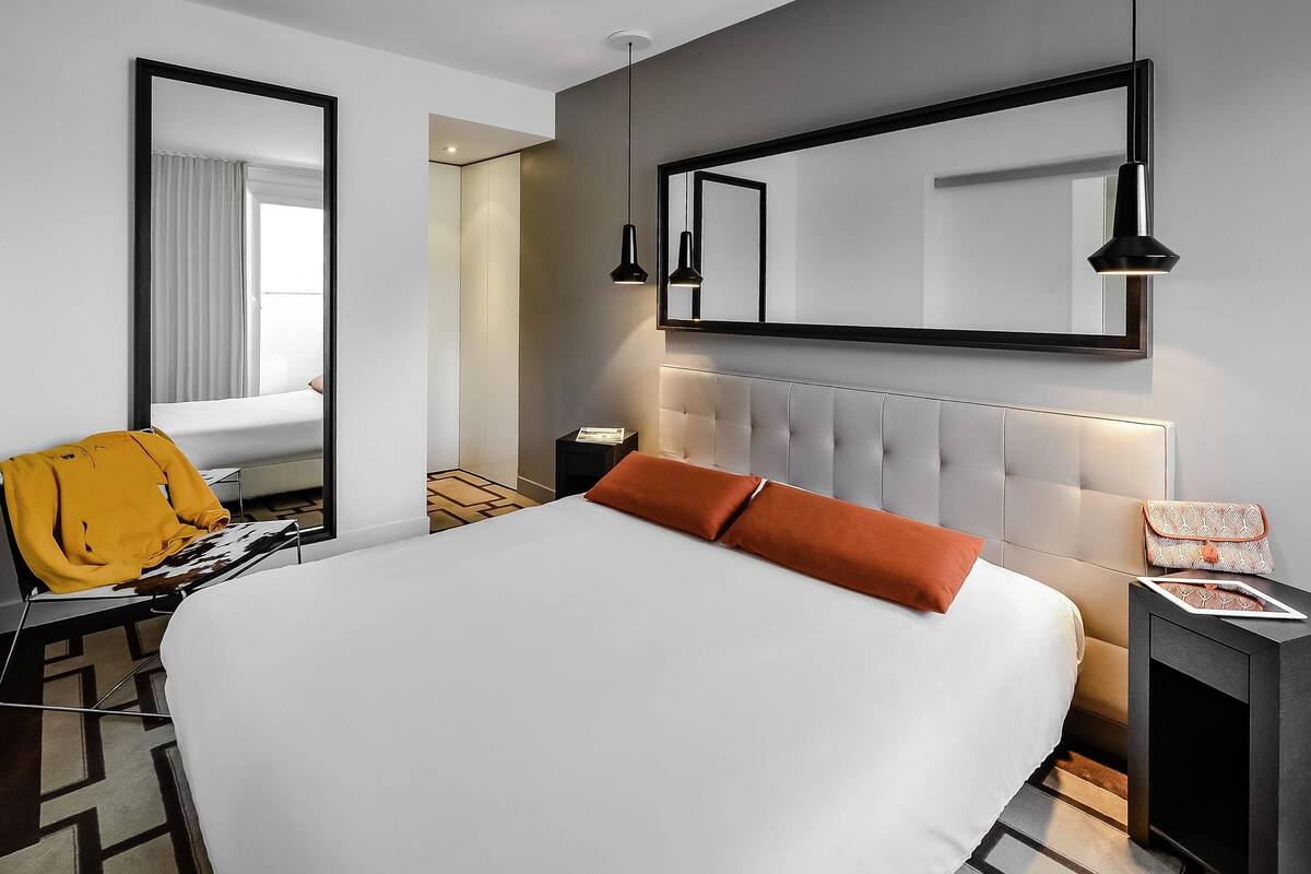 Quarto de hotel moderno com uma cama grande, uma mesa de cabeceira, uma cadeira, um espelho e um criado-mudo.
