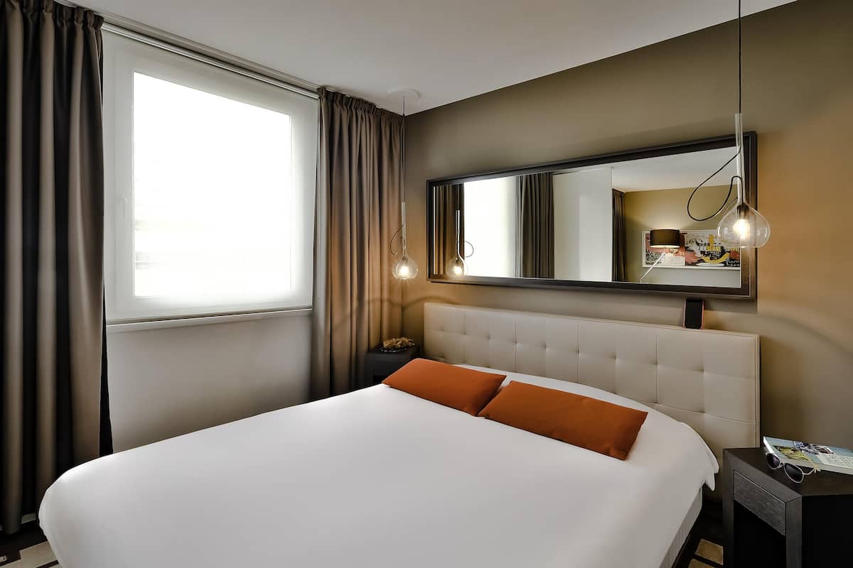 Quarto de hotel com uma cama grande, mesa de cabeceira, espelho e janela com cortinas.