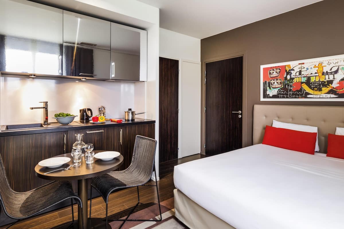 Quarto de hotel compacto com cozinha americana, área de refeições com mesa redonda e cadeiras, cama com almofadas vermelhas e uma grande pintura abstrata na parede.
