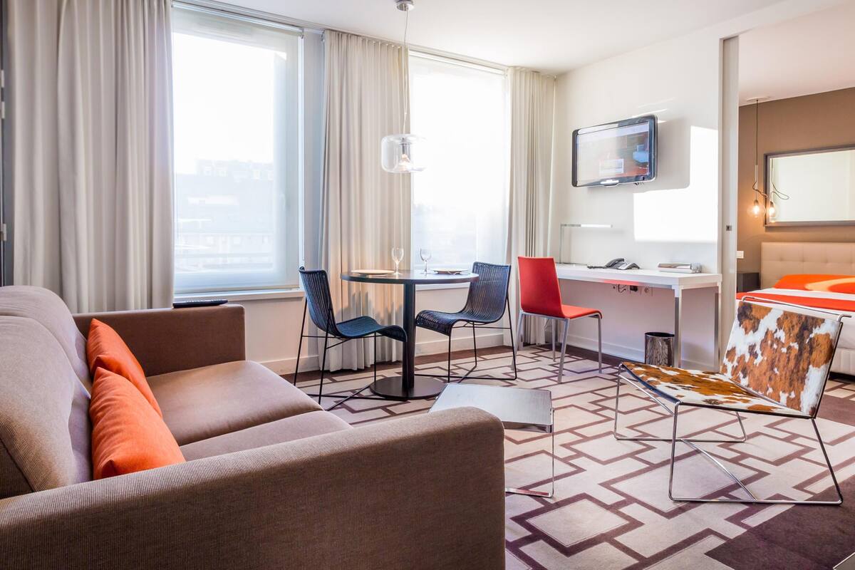Quarto de hotel com sofá, duas cadeiras, mesa pequena, escrivaninha, televisão e cama com cabeceira estampada.
