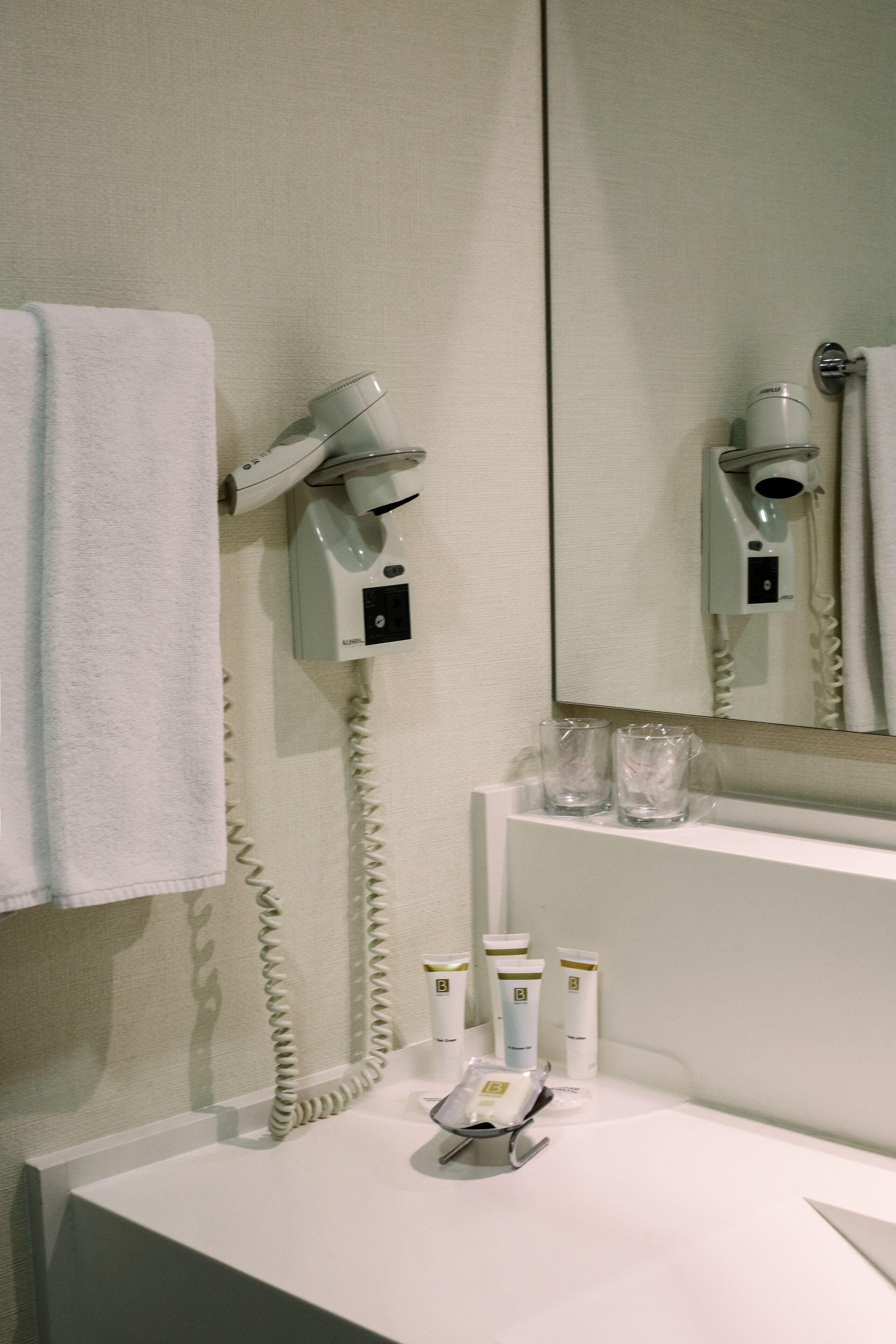 Casa de banho com uma sanita branca, um secador de cabelo fixado na parede, uma toalha pendurada num suporte e um espelho a refletir o secador.