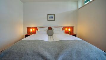 Uma cama de casal com um cobertor cinza, duas mesas de cabeceira com abajures, um quadro na parede e uma janela com persianas.