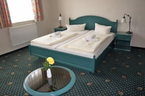 Uma cama de casal com roupa de cama branca, duas mesas de cabeceira com abajures, um vaso com uma flor amarela e um tapete estampado.
