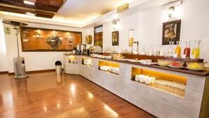 Disposição de buffet com diversos alimentos e bebidas, piso de madeira envernizada e parede decorativa com obras de arte emolduradas.