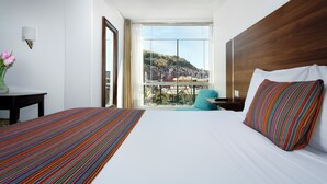 Quarto de hotel com uma cama grande, vista para a cidade, cabeceira de madeira e uma mesinha com um vaso de flores.