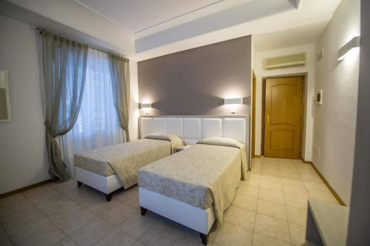 Quarto de hotel com duas camas de solteiro, uma janela grande com cortinas, uma porta de madeira e um ar condicionado fixado na parede.