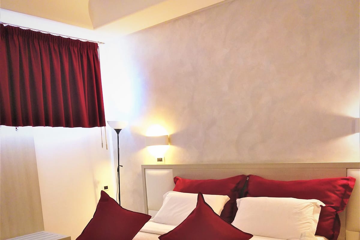 Uma cama bem arrumada com roupa de cama branca e almofadas vermelhas, uma mesa de cabeceira com controle remoto e um abajur na parede.